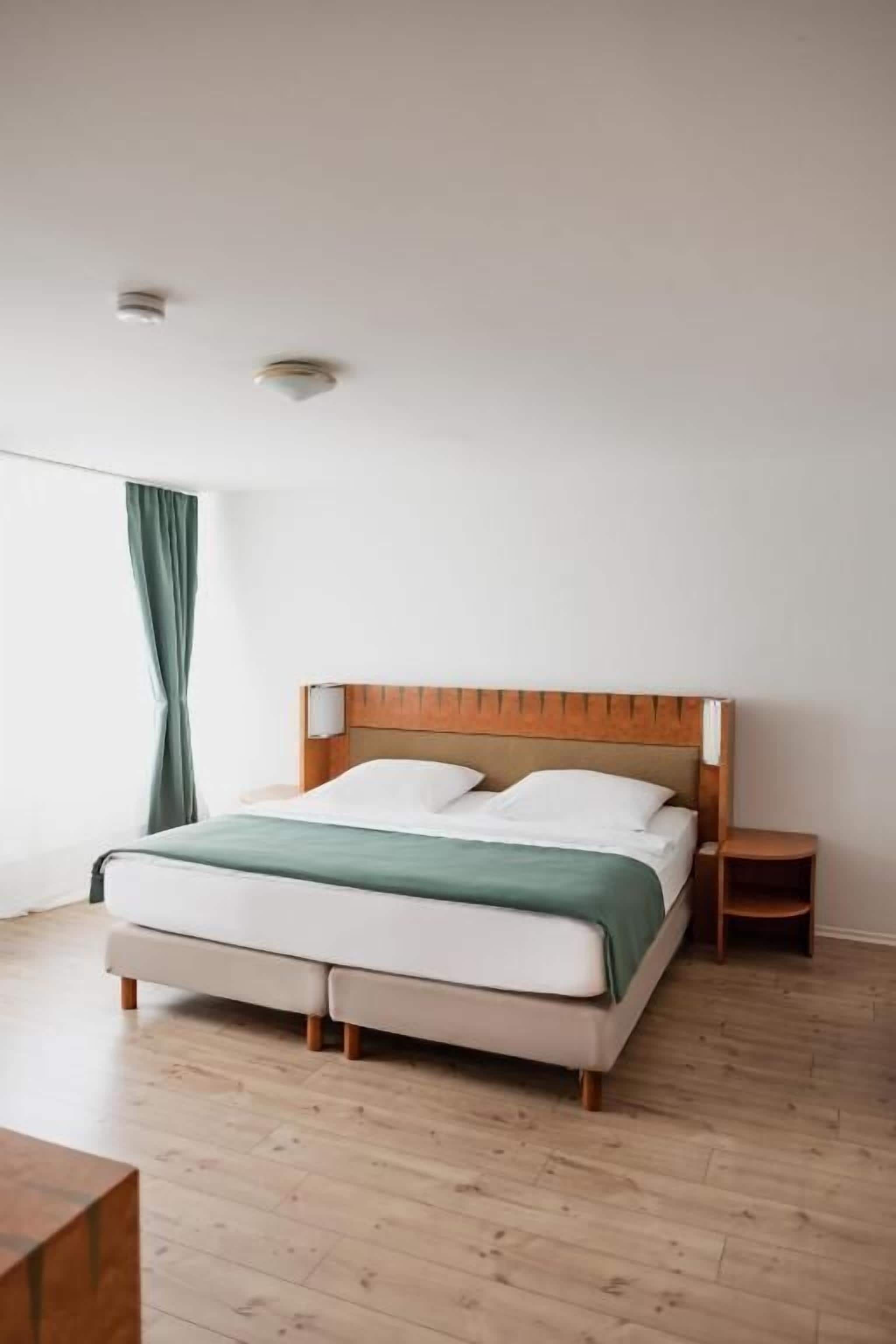 Quarto de hotel moderno com cabeceira de madeira, uma cama de solteiro, mesa de cabeceira e cortina verde.