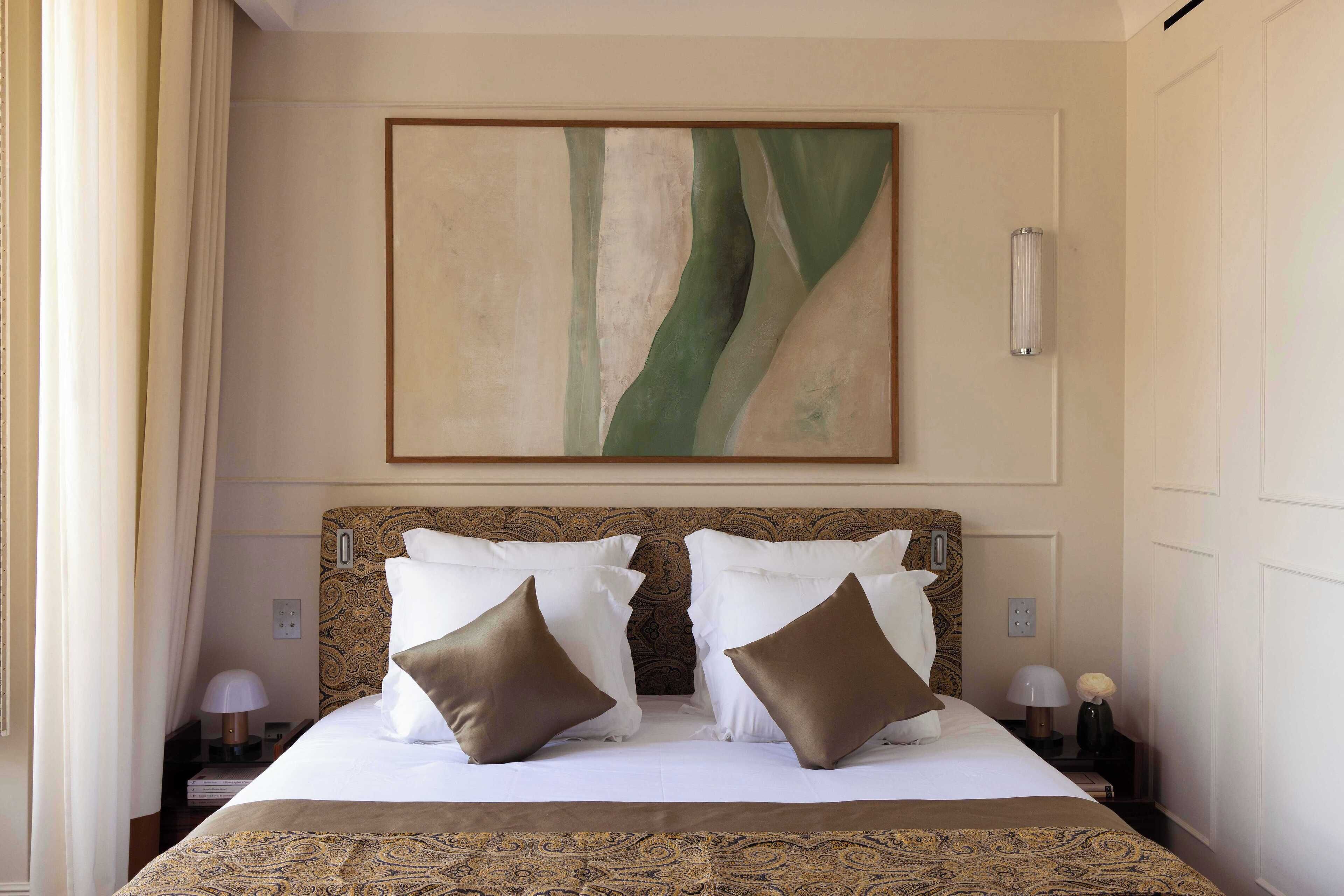 Uma cama bem arrumada com cabeceira estampada, uma grande pintura abstrata acima e uma mesa de cabeceira com abajur à direita.