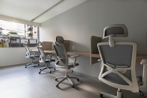 Um espaço de escritório moderno com quatro cadeiras de escritório e uma mesa, janelas amplas com vista para edifícios e árvores, e um design limpo e minimalista.