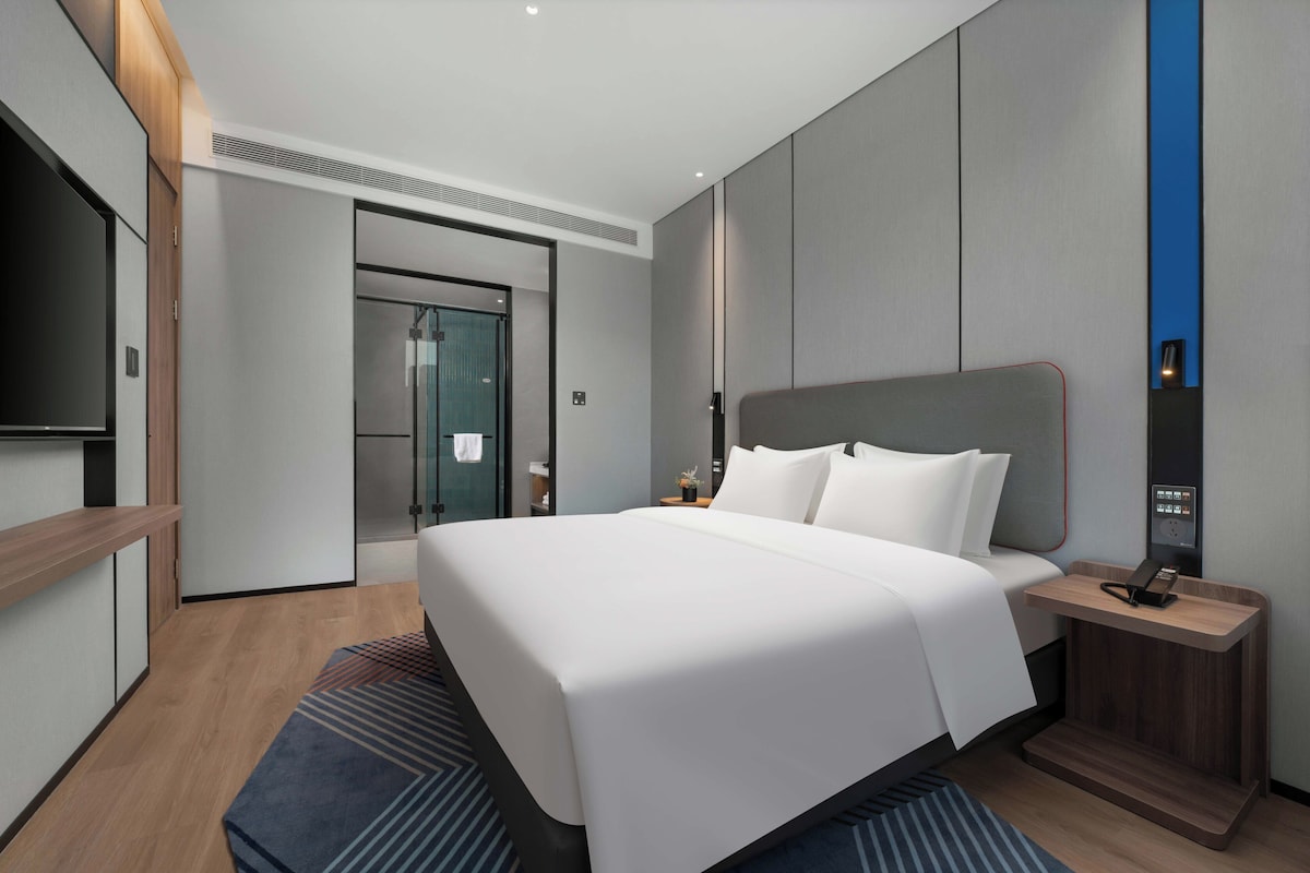 Quarto de hotel moderno com cama grande, mesa de cabeceira com telefone, TV integrada e porta de vidro para o banheiro.