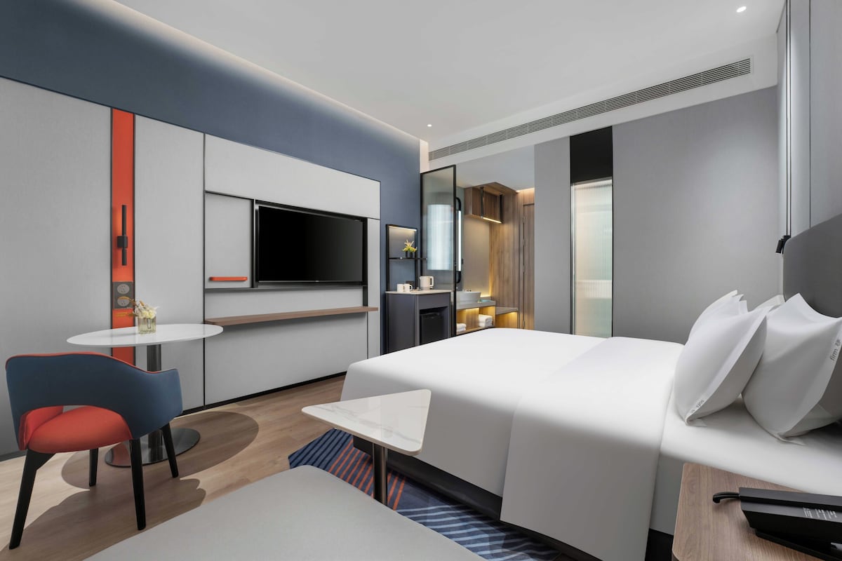 Quarto de hotel moderno com uma cama, uma pequena escrivaninha, uma cadeira, televisão e minibar.