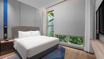 Quarto de hotel moderno com uma cama grande, mesa de cabeceira, TV e janela com vista para vegetação.