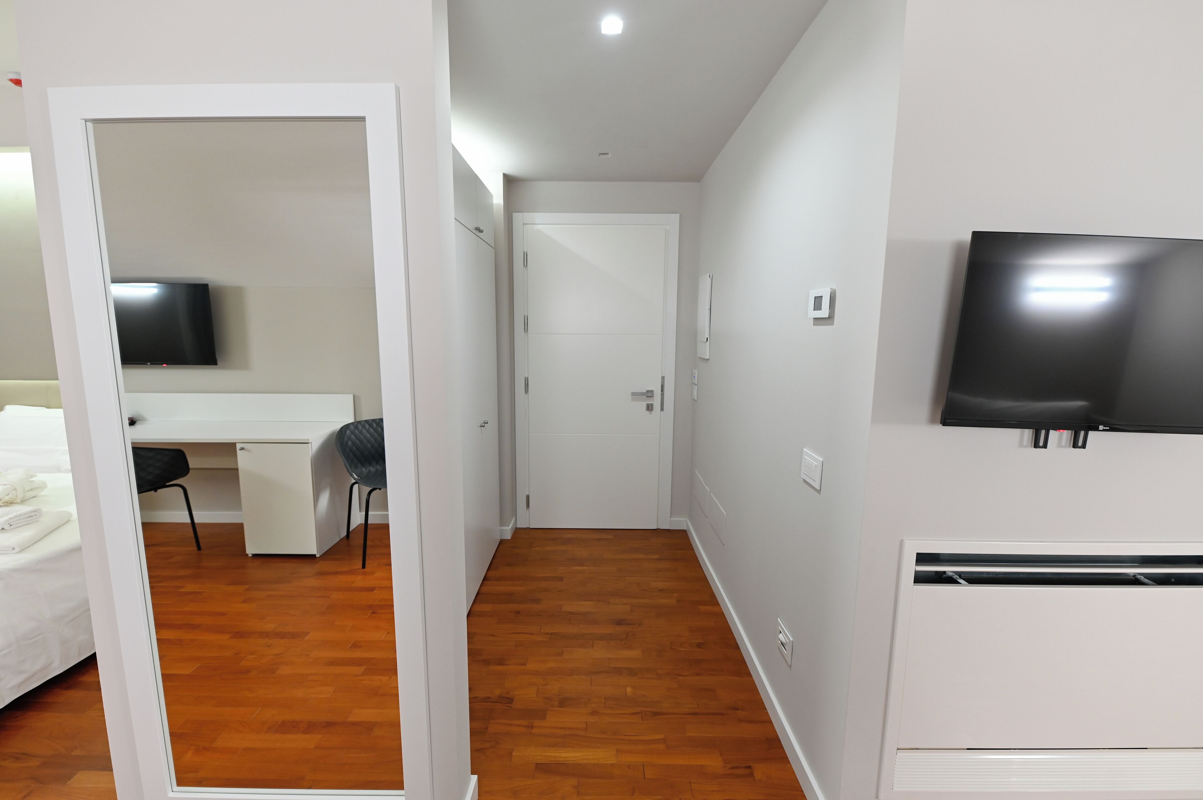 Quarto de hotel com piso de madeira, paredes brancas, uma cama, uma escrivaninha com cadeira, televisão e uma porta.