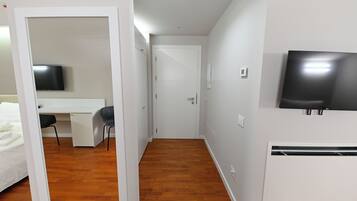 Quarto de hotel com piso de madeira, paredes brancas, uma cama, uma escrivaninha com cadeira, televisão e uma porta.