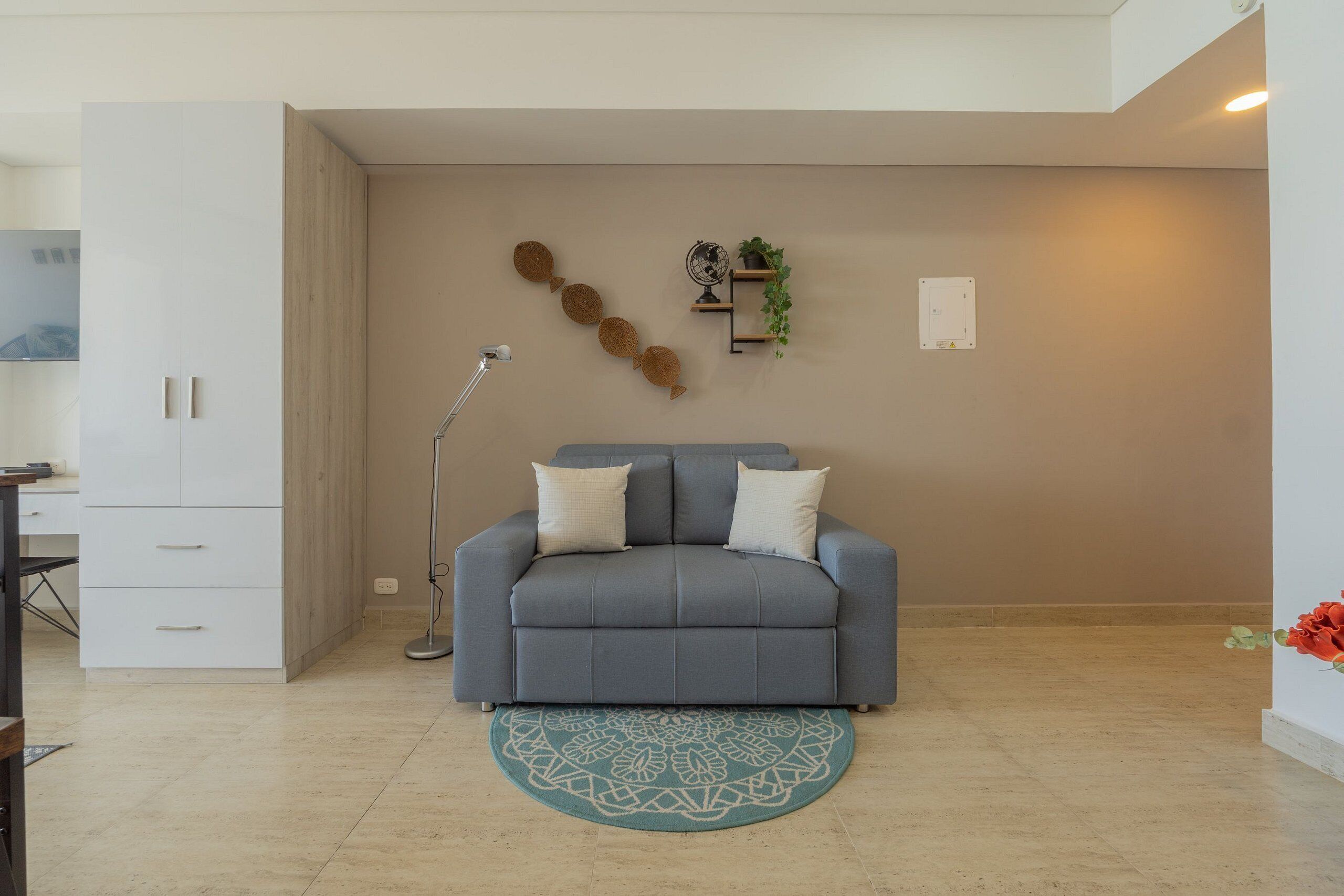 Uma sala de estar moderna com um sofá cinza, um tapete redondo estampado, uma luminária de chão, um armário branco e uma prateleira fixada na parede com uma planta.