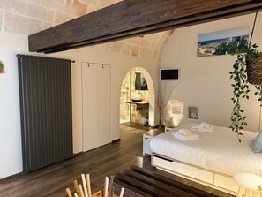 Quarto com cama, piso de madeira, parede de pedra e vista para um banheiro com pia e chuveiro.