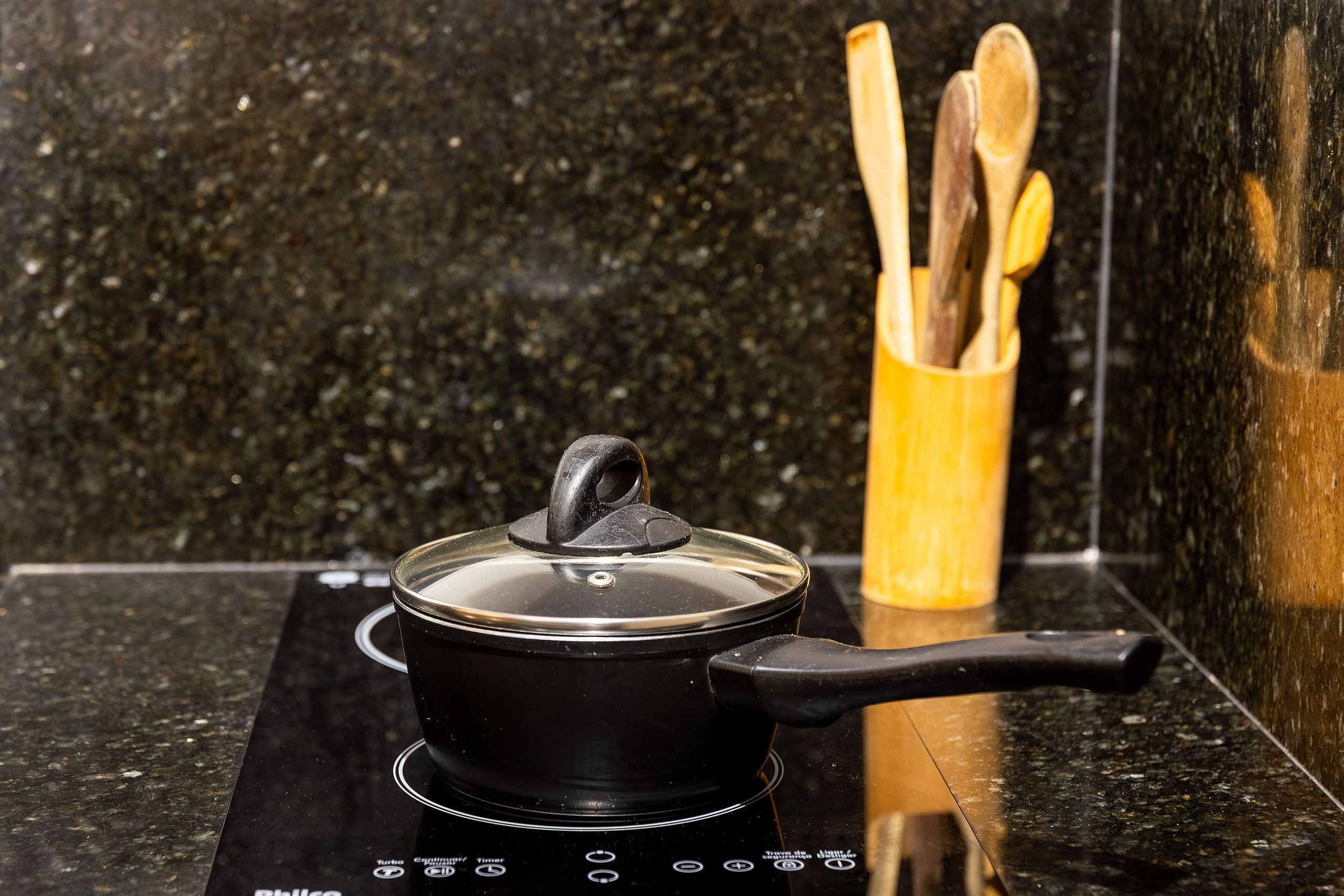 Um fogão elétrico preto com uma panela em cima, um porta-utensílios de madeira e uma bancada de granito.