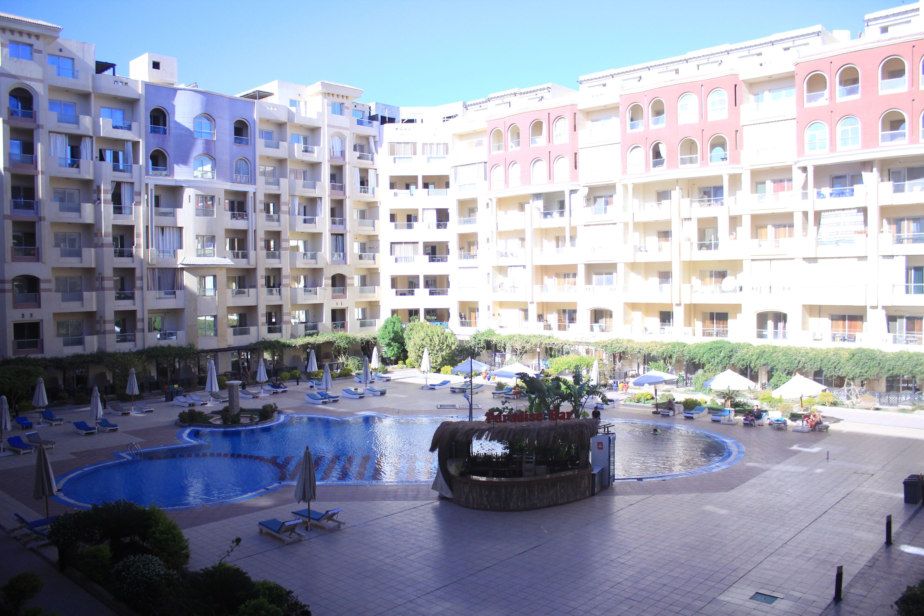 Pátio de hotel com piscina central, espreguiçadeiras e guarda-sóis.