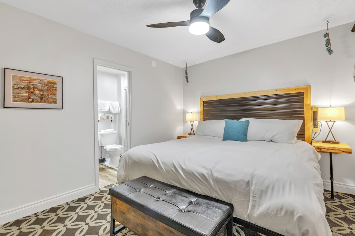 Quarto com cama, um banquinho, ventilador de teto, mesa de cabeceira com abajur e quadro na parede.