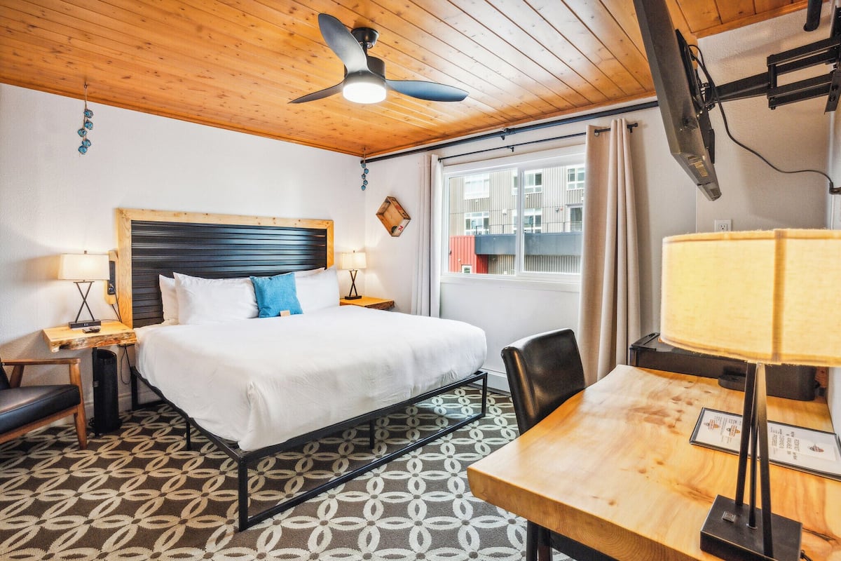 Quarto de hotel com cama, escrivaninha, cadeira, ventoinha no teto, janela e vista para edifícios.
