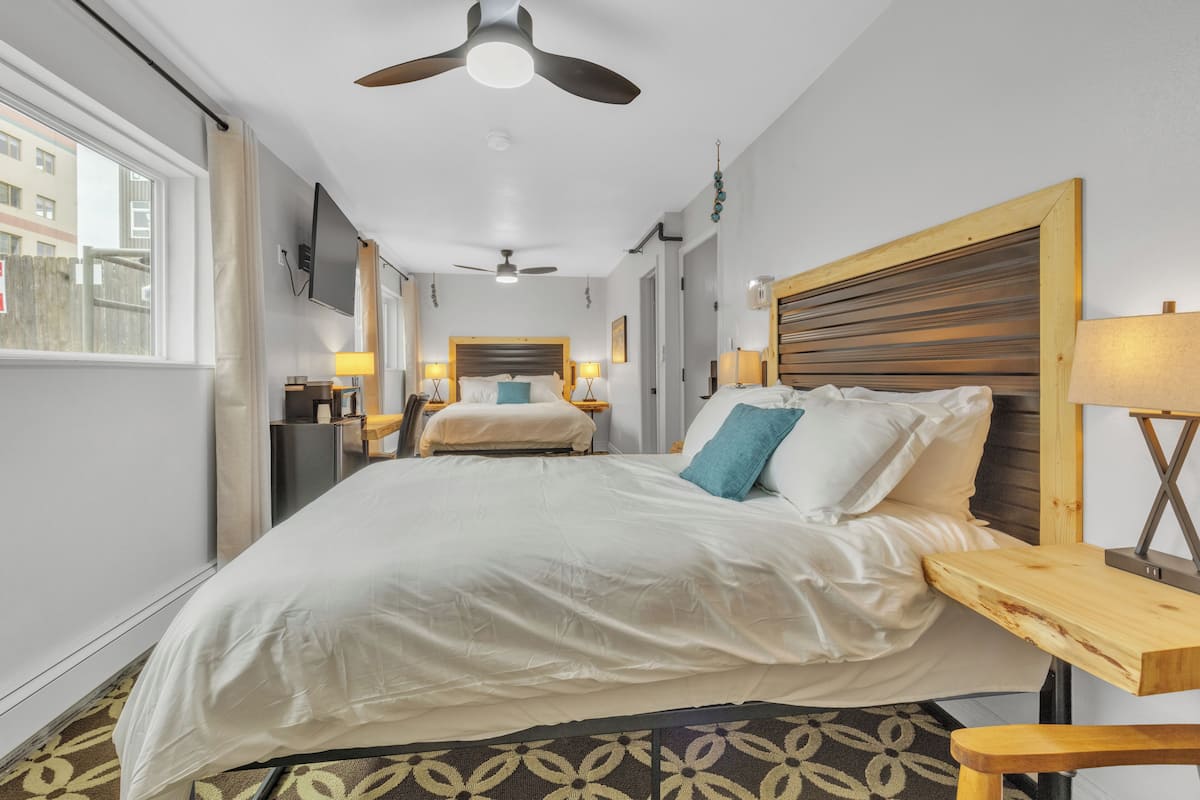 Quarto de hotel com uma cama grande, cabeceira de madeira, escrivaninha com luminária e ventilador de teto.