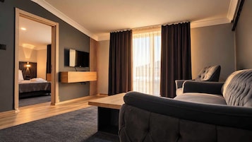Quarto de hotel moderno com sofá, televisão de tela plana na parede, mesa lateral de madeira e uma janela grande com cortinas.