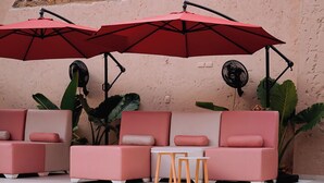 Uma área de estar à beira da piscina com sofás cor-de-rosa e duas mesas de madeira, rodeada por grandes guarda-sóis vermelhos.