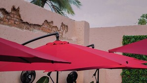 Área de um hotel com espaços de estar ao ar livre, incluindo sofás rosa e brancos, mesas redondas e guarda-sóis vermelhos para proteção contra o sol.