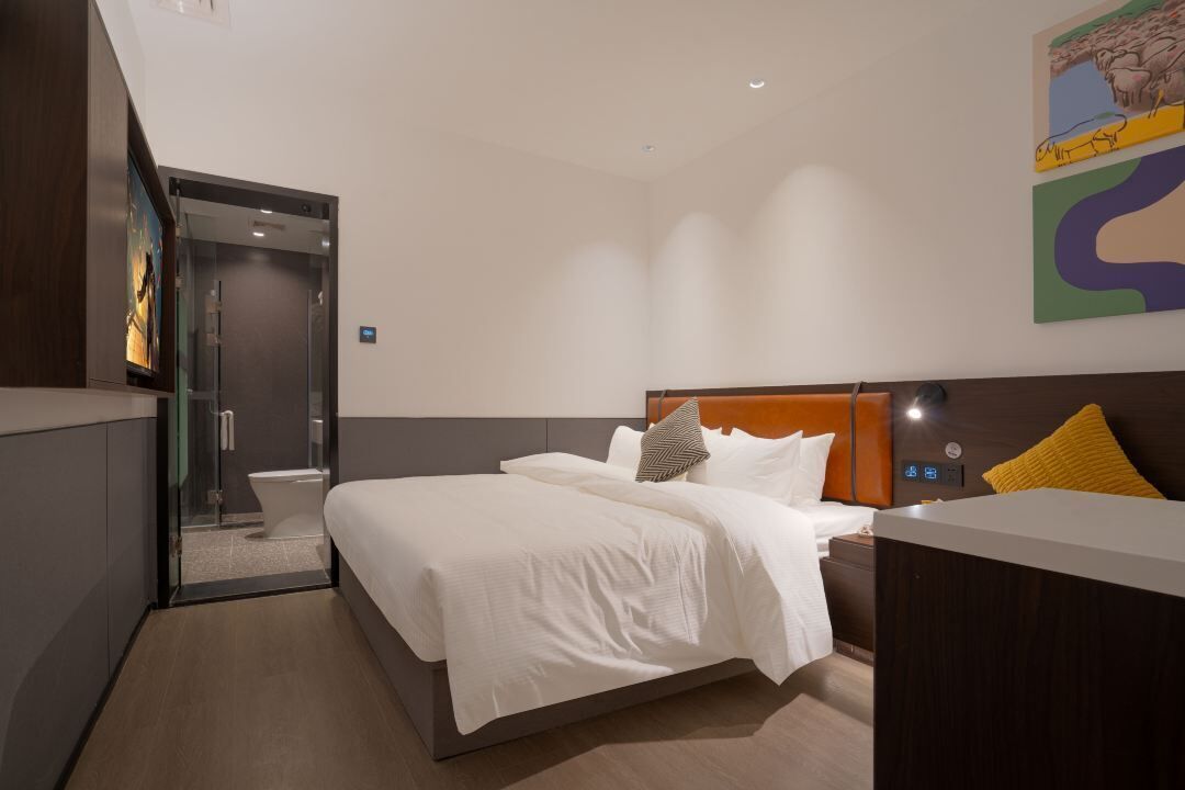 Quarto de hotel moderno com uma cama grande, cabeceira de madeira e área de chuveiro com divisória de vidro.