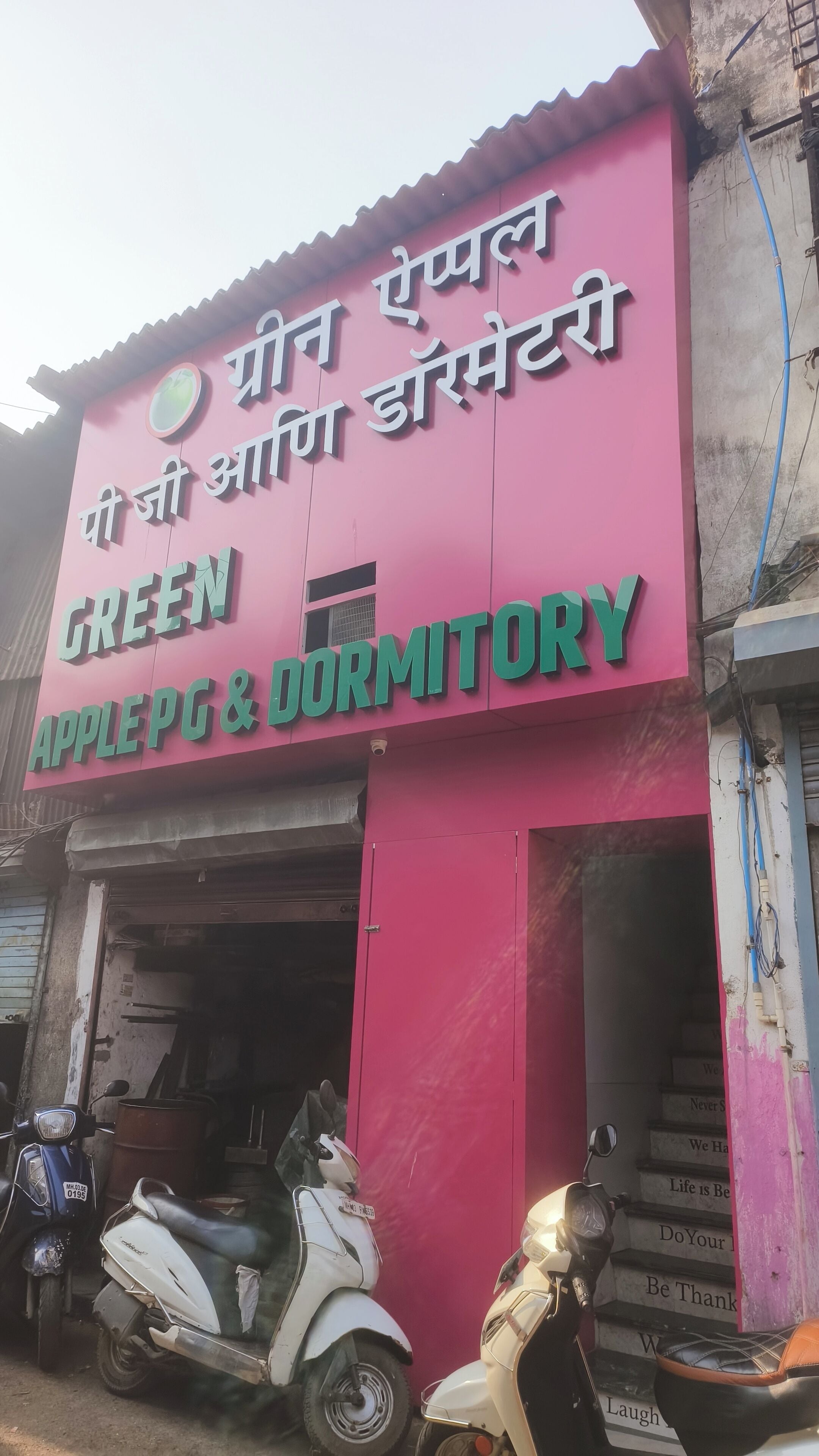 Um edifício com uma placa que diz "GREEN APPLEPG & DORMITORY" em inglês e outro idioma. Duas scooters estão estacionadas na frente.