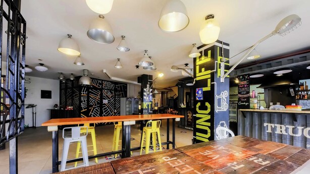 Interior moderno com mesa de madeira, banquetas amarelas e um espaço bar com letreiro neon.