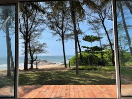 Vista de um quarto com grandes janelas para uma praia com palmeiras e um deck de madeira.