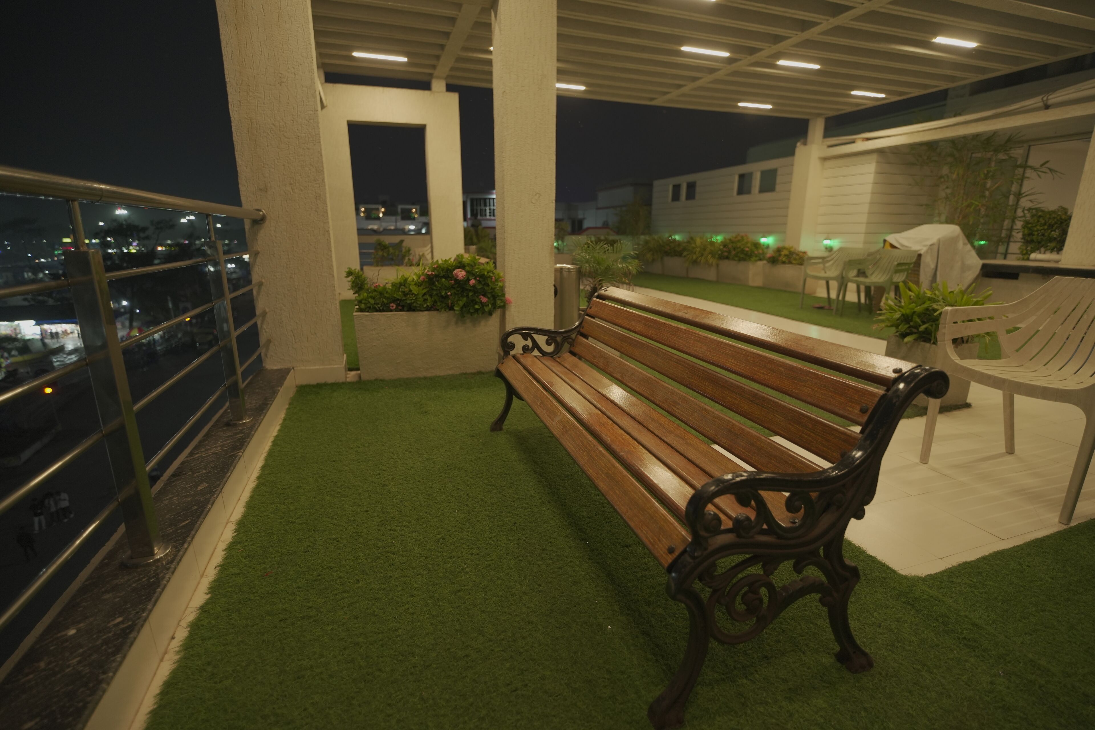 Uma varanda com um banco de madeira, plantas em vasos e vista da cidade à noite.