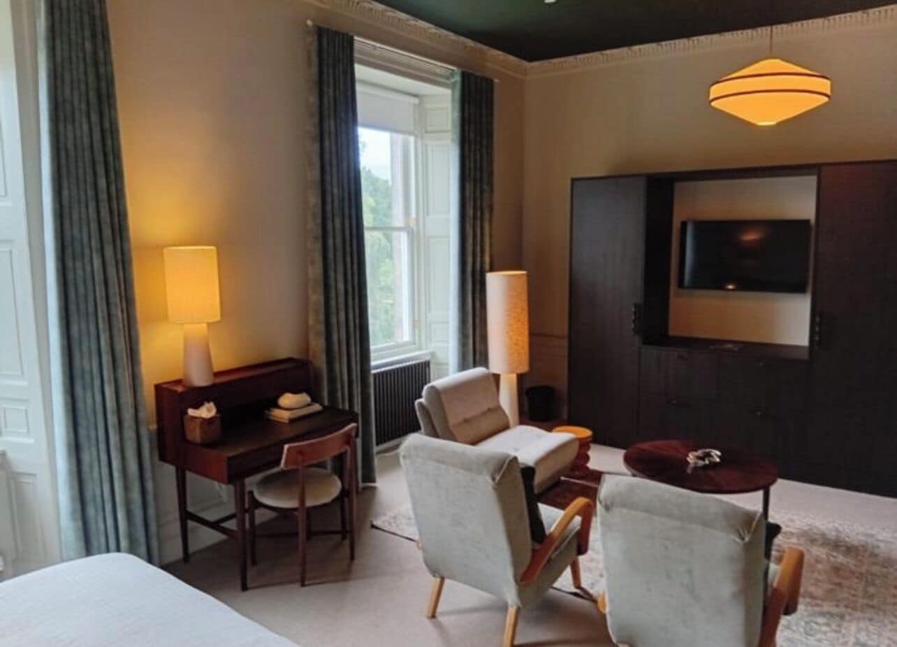 Quarto de hotel com uma cama, duas poltronas, uma escrivaninha com abajur, uma mesinha redonda, televisão na parede e uma janela com cortinas.