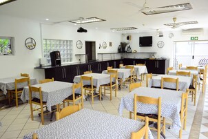 Uma sala de jantar com várias mesas e cadeiras, uma cozinha ao fundo e um ventilador de teto.
