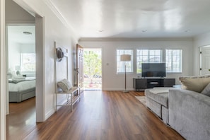 Uma sala de estar espaçosa com piso de madeira, um sofá cinza, um suporte para TV e uma porta que dá para uma varanda.