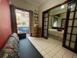 Quarto com sofá, mesa pequena, espelho e porta de correr para uma varanda.
