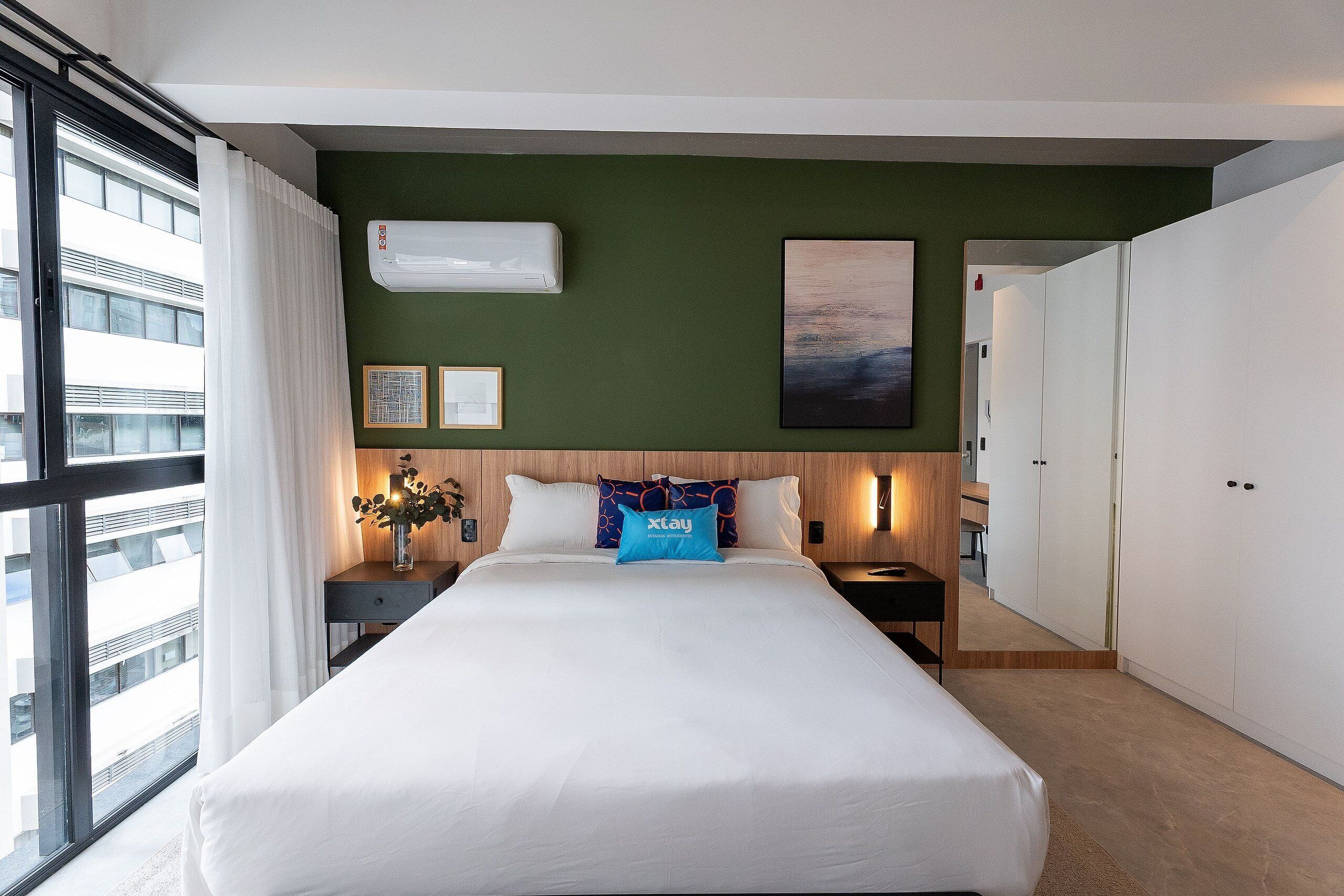 Quarto de hotel moderno com uma cama grande, uma mesa de cabeceira, ar condicionado, um quadro emoldurado e vista da cidade por uma grande janela.