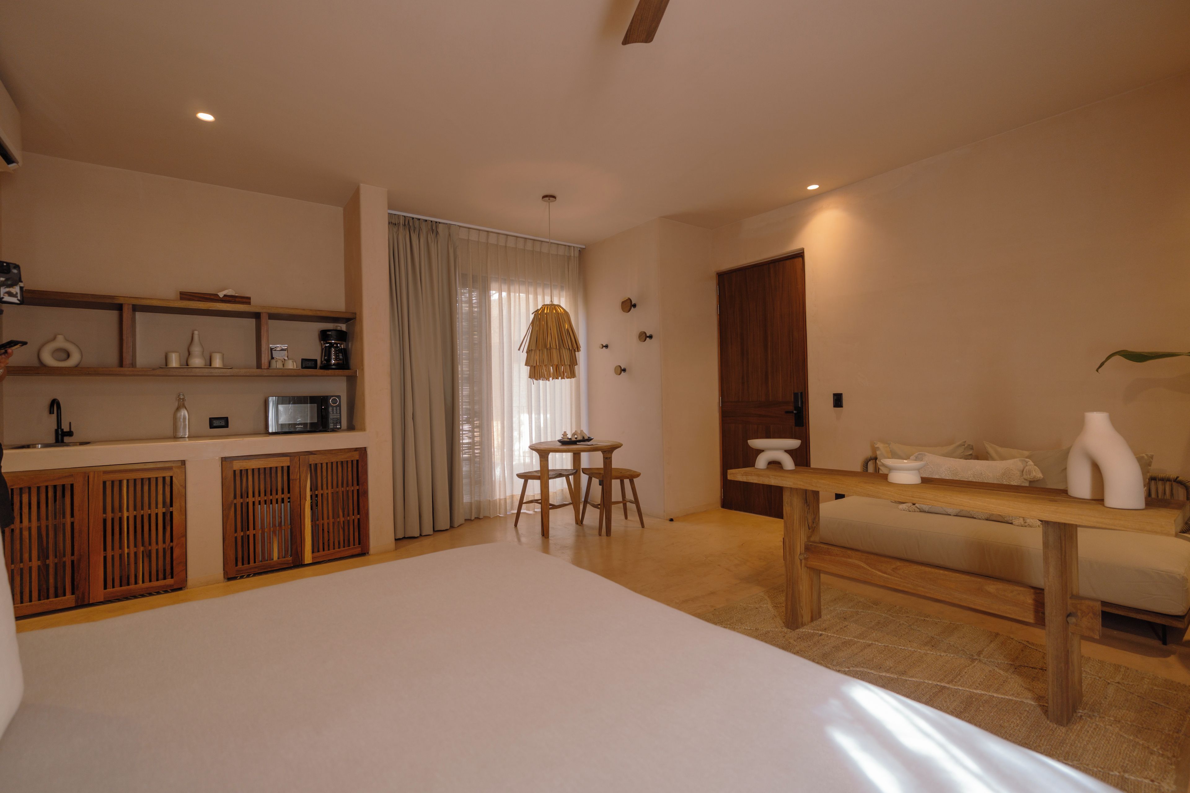 Quarto moderno e bem iluminado com cama, área de refeições com mesa e cadeiras, cozinha equipada com micro-ondas e cafeteira, e porta de madeira.