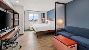 Quarto de hotel com uma cama, uma escrivaninha com computador, televisão, sofá e uma mesinha.