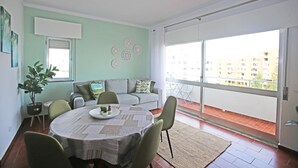 Sala de estar com sofá, mesa de jantar, cadeiras, uma planta em vaso e uma varanda com vista para edifícios.