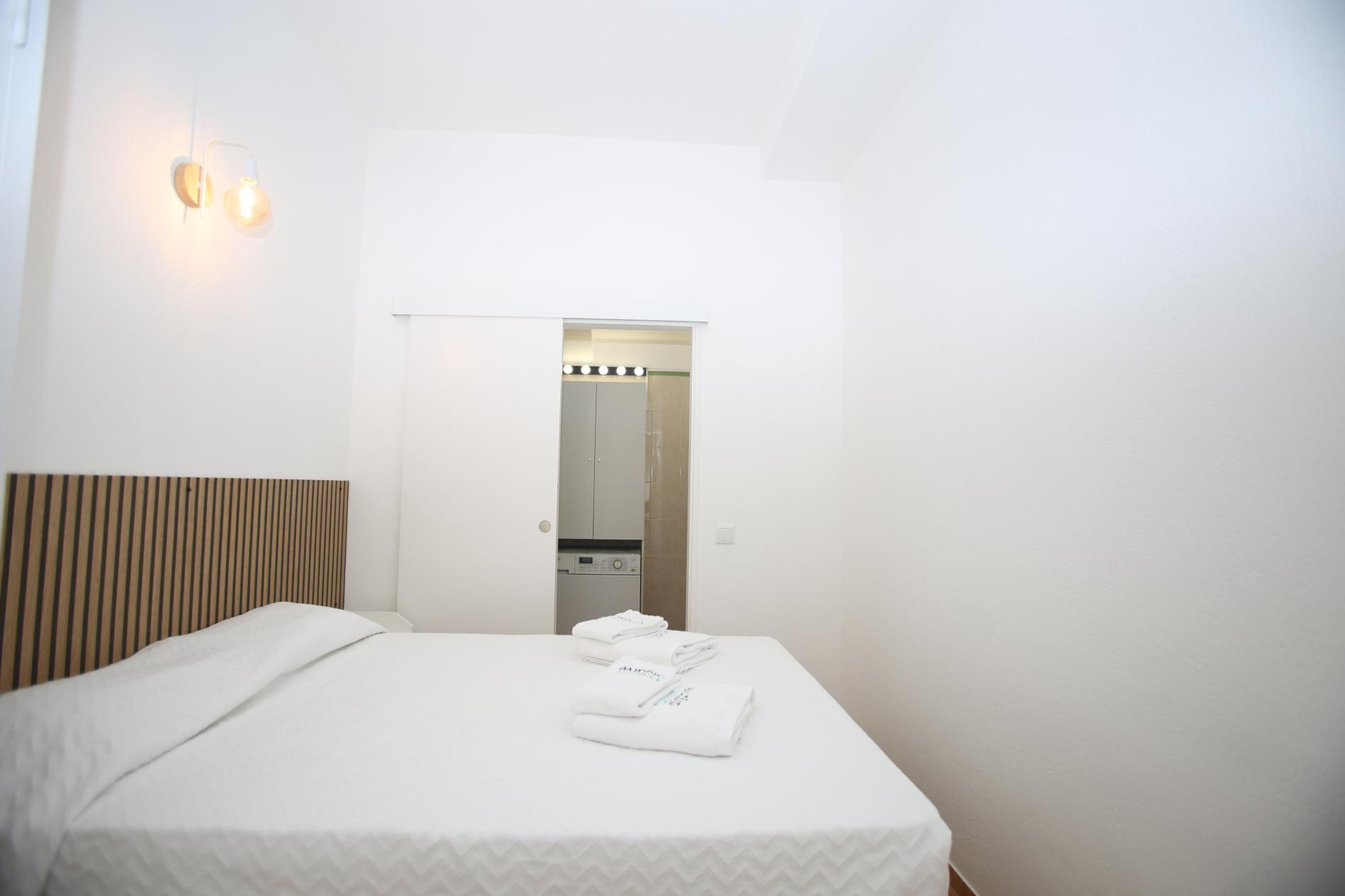 Quarto de hotel com cama, cabeceira de madeira, luminária fixa na parede e espelho iluminado acima de uma porta.