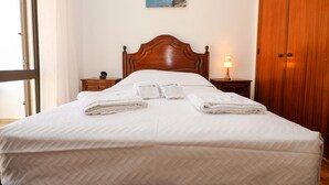 Cama bem arrumada com toalhas dobradas, cabeceira de madeira, mesa de cabeceira com abajur e quadro com uma paisagem costeira na parede.