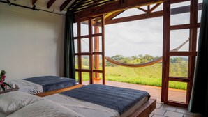 Quarto com duas camas, uma rede e vista para uma paisagem verde através de grandes janelas.