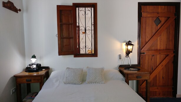 Quarto com cama, duas mesas de cabeceira, porta de madeira, janela com grade decorativa e telefone fixado na parede.