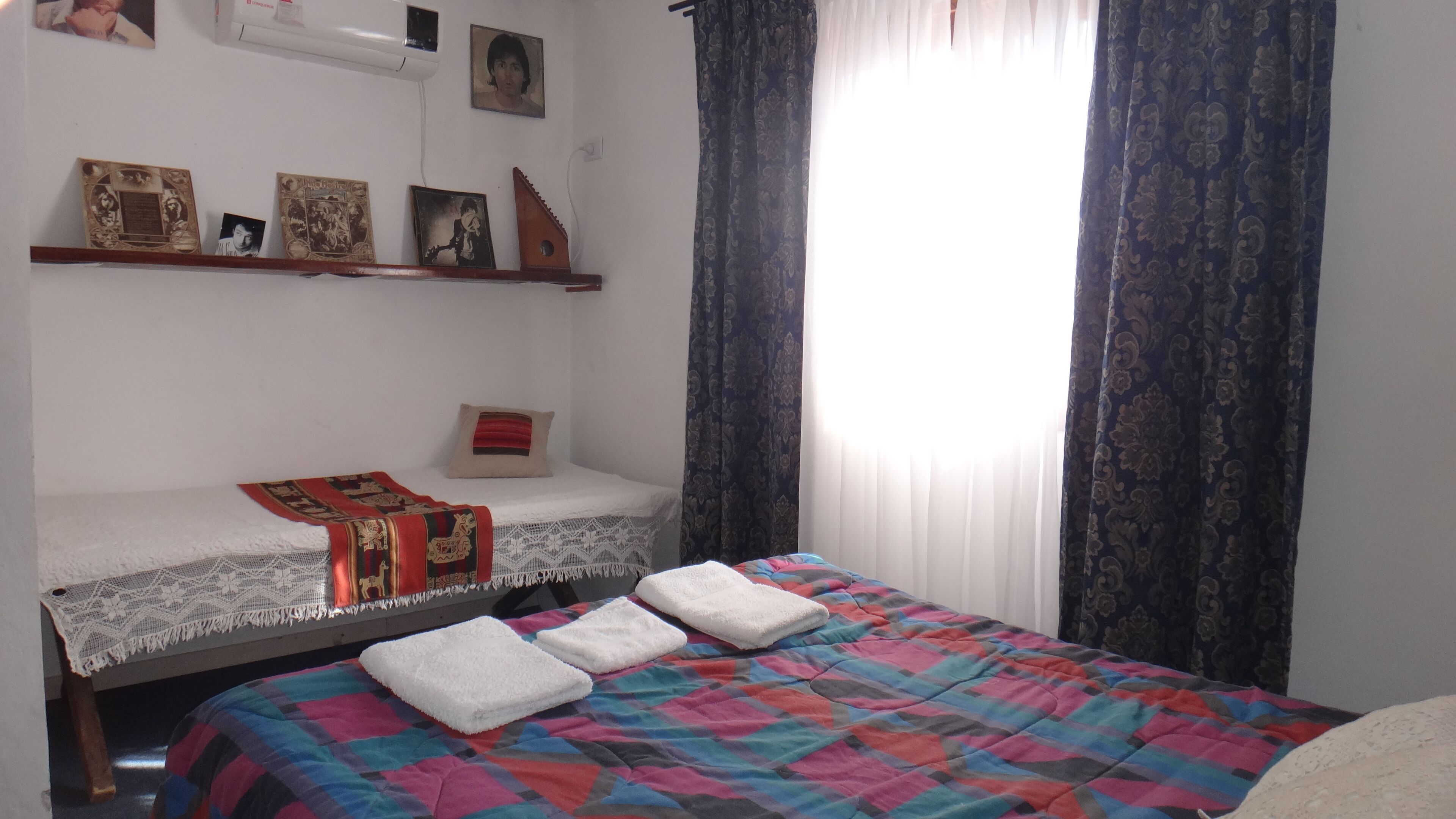 Quarto com cama, mesa de cabeceira, prateleira na parede com fotos e janela com cortinas.