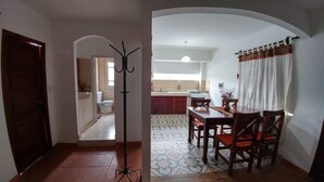 Um corredor que dá para uma sala de jantar com piso ladrilhado, uma mesa com cadeiras e uma cozinha ao fundo.