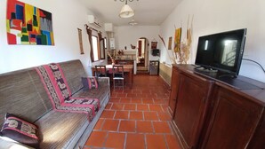 Uma sala de estar com sofá, televisão sobre um móvel de madeira, e cozinha ao fundo.