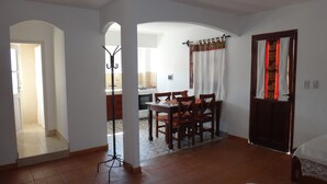 Quarto com uma mesa de jantar e cadeiras, cozinha ao fundo e uma porta com cortinas.