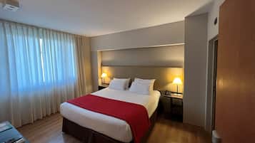 Quarto de hotel com uma cama grande, duas luminárias de cabeceira, um cobertor vermelho, piso de madeira e uma janela com cortinas.
