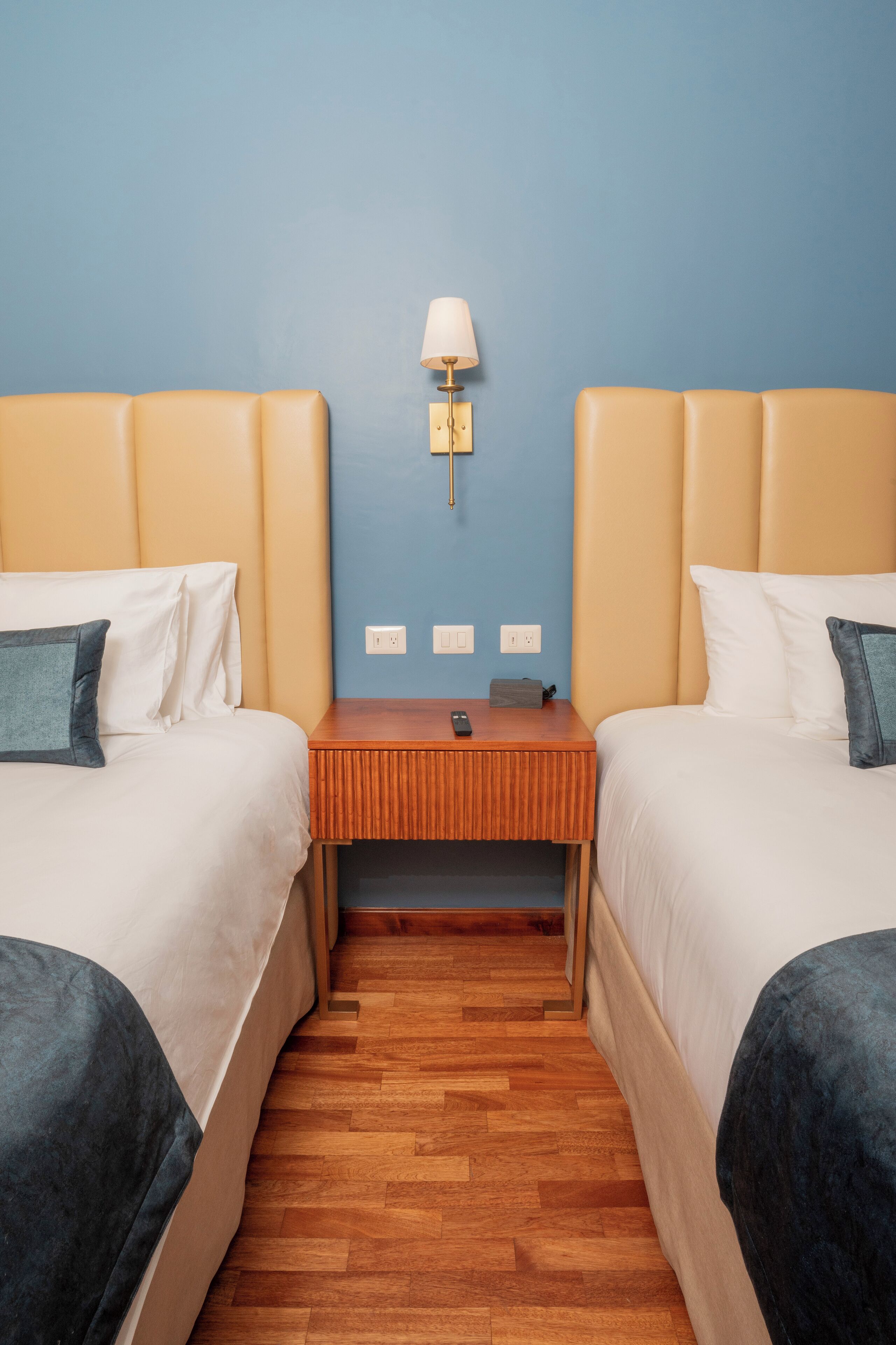 Quarto de hotel com duas camas, uma mesa de cabeceira de madeira, uma lâmpada e uma tomada na parede.