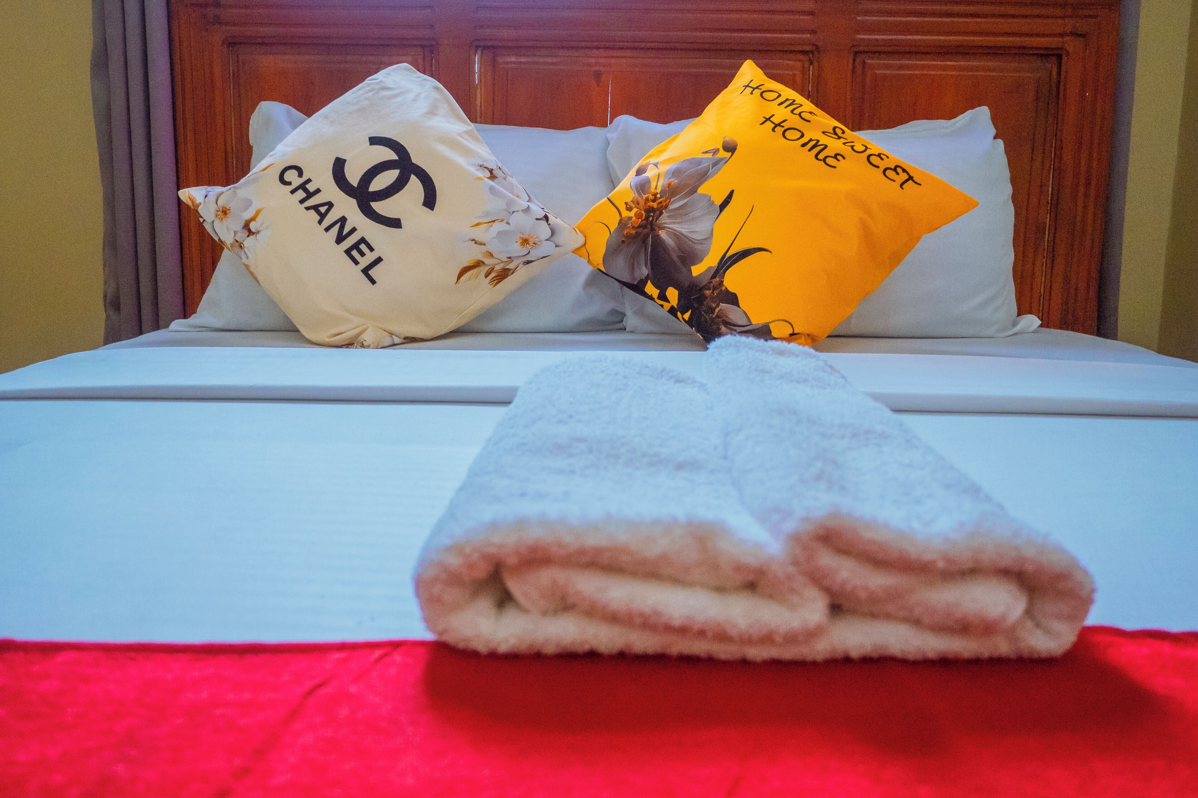 Uma cama bem arrumada com uma toalha dobrada, dois travesseiros com estampas florais e um cobertor vermelho.