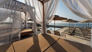Um espaço de lounge à beira-mar com assentos acolchoados e guarda-sóis, com vista para uma piscina e o mar.