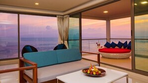 Quarto moderno com uma grande janela, um sofá azul, uma mesa branca com frutas e um assento tecido em vermelho e branco.