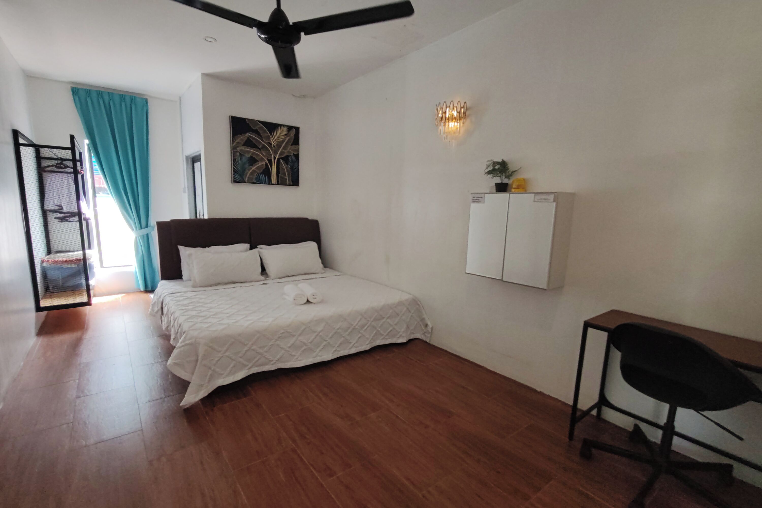 Quarto com cama, escrivaninha, ventoinha no teto, guarda-roupa e um quadro na parede.
