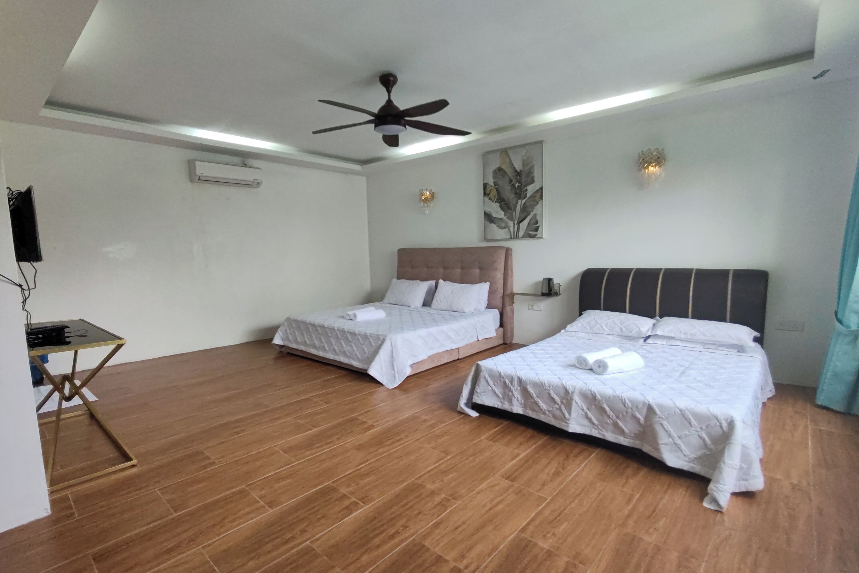 Quarto de hotel com duas camas, um ventilador de teto, um ar condicionado, um quadro e uma pequena mesa com abajur.
