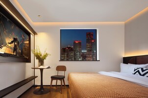 Quarto de hotel moderno com vista para a cidade, uma cama com colcha estampada, uma pequena mesa com uma cadeira e um quadro na parede.