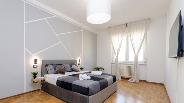 Quarto moderno com uma cama grande, uma mesinha de cabeceira com luminária, uma planta em vaso e uma janela com cortinas.