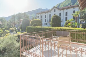 Uma varanda com grades de ferro forjado, área de estar com mesa e cadeiras, e vista para um edifício branco com várias janelas e uma varanda.