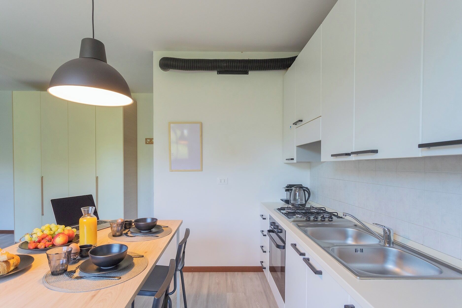 Uma cozinha moderna com armários brancos, fogão a gás e uma pia. Há uma mesa de jantar com itens de café da manhã e uma luminária pendente sobre ela.